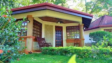 Um edifício de dois andares com telhado vermelho, varanda coberta e um jardim com diversas plantas e um banco.