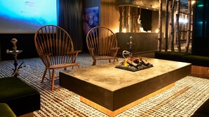 Um hall de hotel moderno com uma tela grande exibindo 'HOTEL KUMO', duas cadeiras de madeira, uma mesa de centro com lareira e um balcão de recepção.