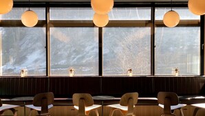 Restaurante com interior moderno, cadeiras de madeira, chão quadriculado e grandes janelas com vista para uma paisagem nevada.