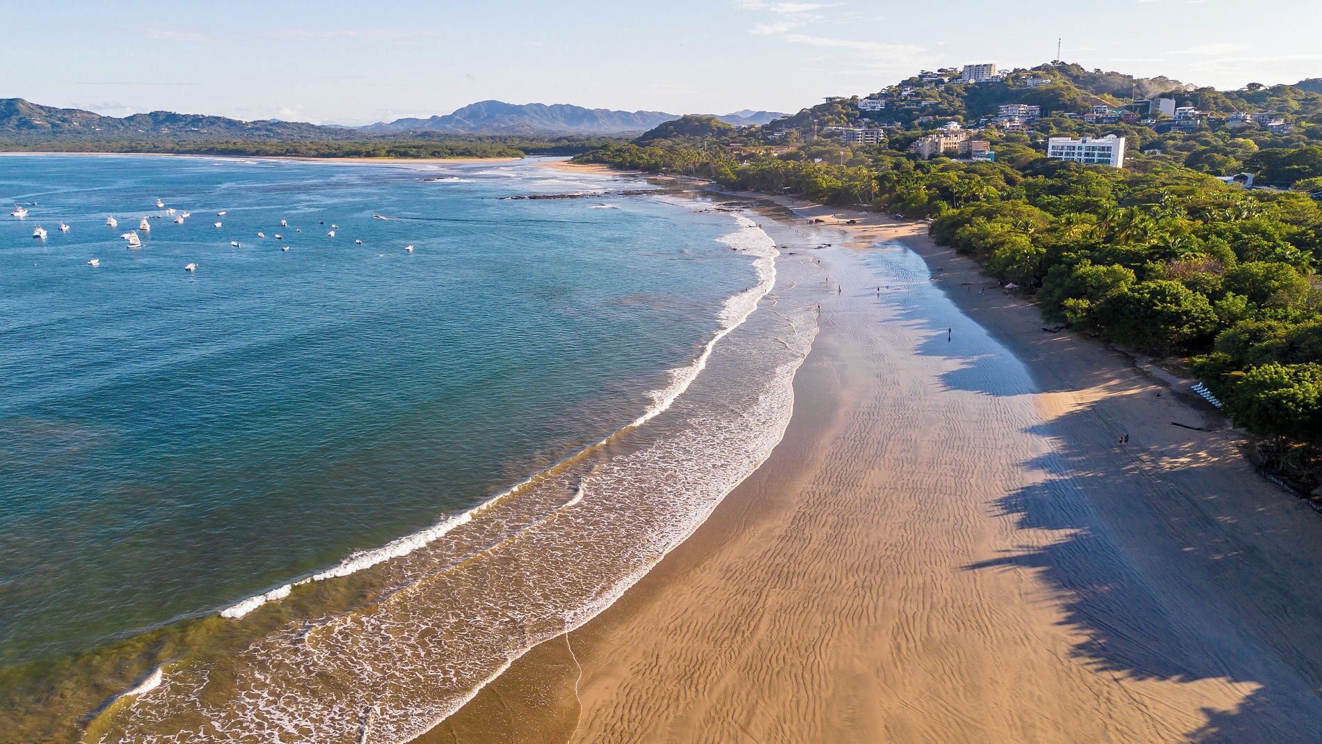 Vista aérea de uma praia com águas cristalinas, areal arenoso e edifícios ao fundo.