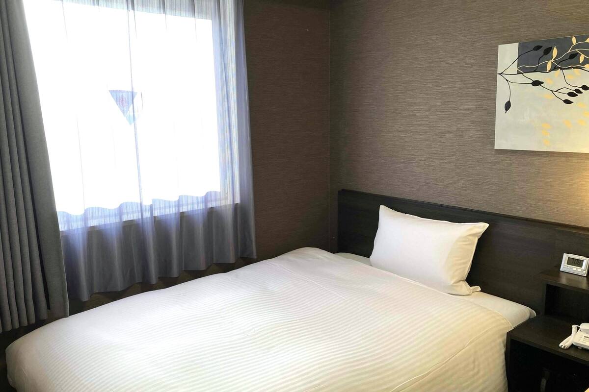 Quarto de hotel com uma cama grande, mesa de cabeceira com telefone e janela com cortinas leves.