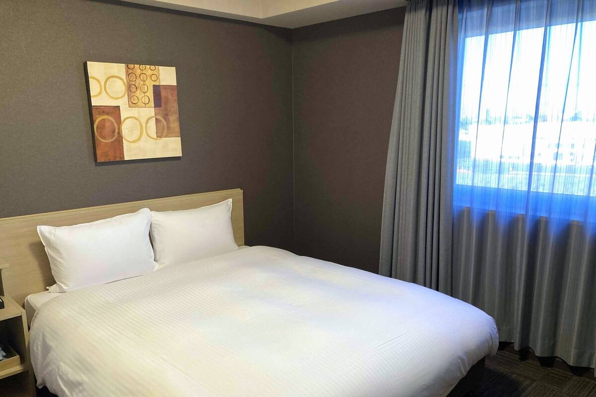 Quarto de hotel com uma cama grande, cabeceira, mesa de cabeceira e uma janela com cortinas.