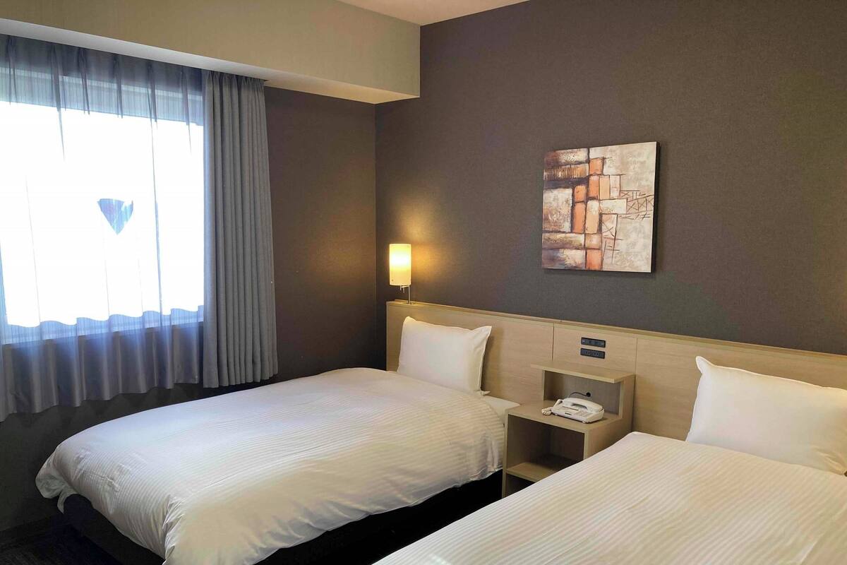 Quarto de hotel com duas camas, uma mesa de cabeceira com telefone, uma luminária na parede e um quadro.