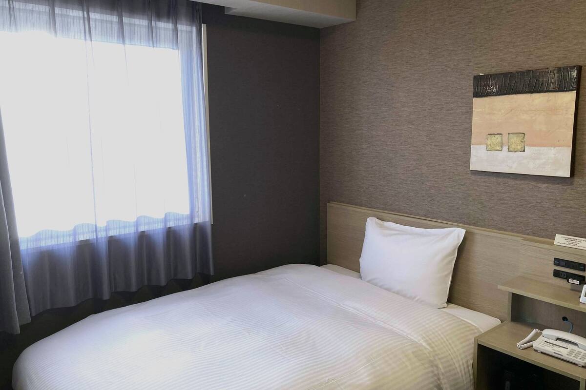 Quarto de hotel com uma cama grande, uma mesa de cabeceira com telefone e luminária, uma janela com cortinas leves e uma obra de arte na parede.