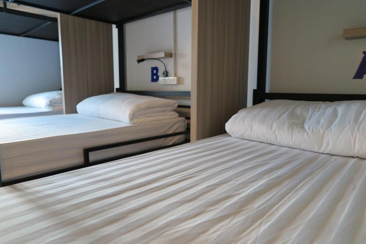 Quarto de hotel com duas camas, cabeceira de madeira, luminária na parede e decoração com a letra 'B'.
