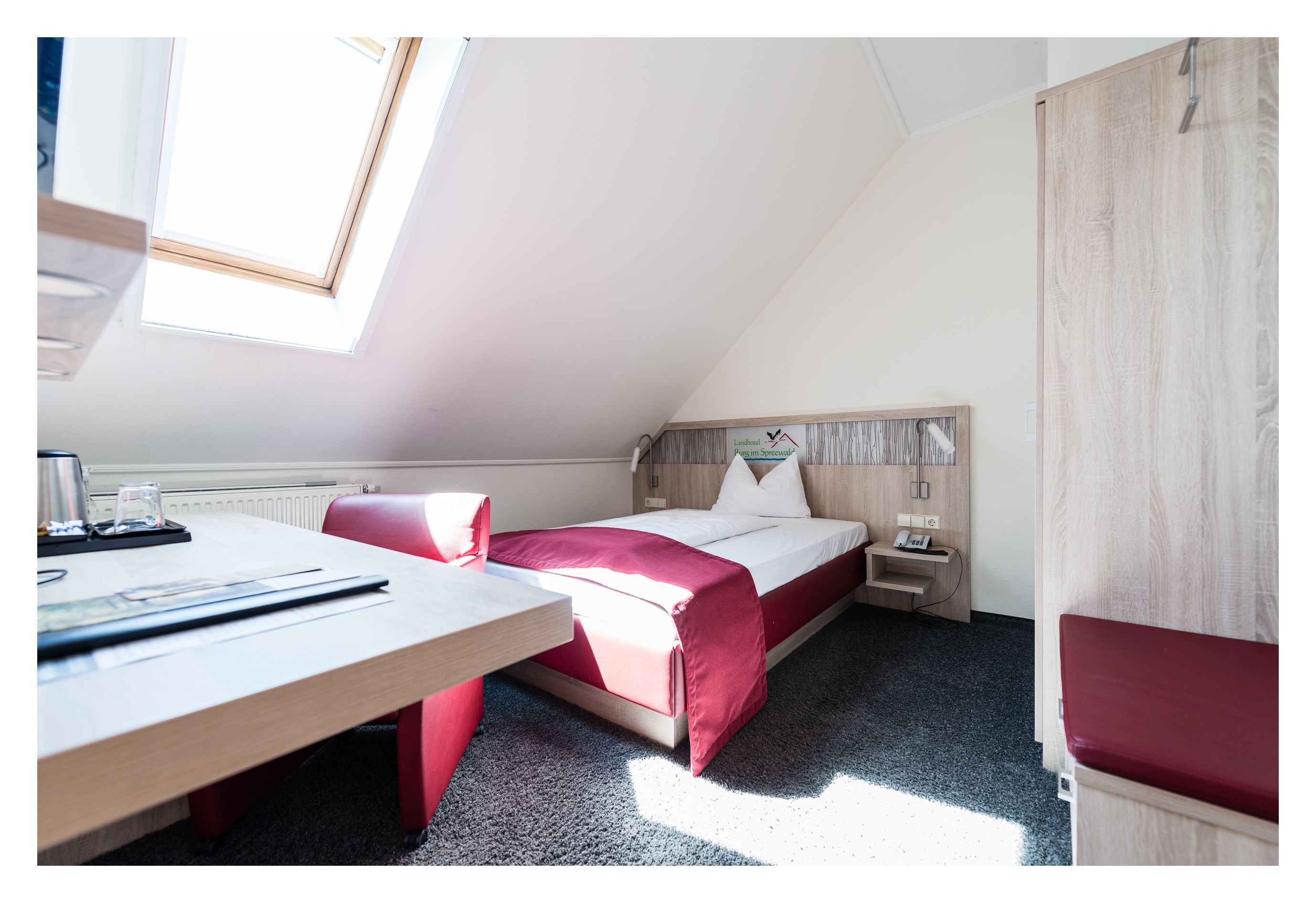 Quarto moderno e bem iluminado, com teto inclinado, cama com roupa de cama vermelha, escrivaninha de madeira e pequena mesa de cabeceira.
