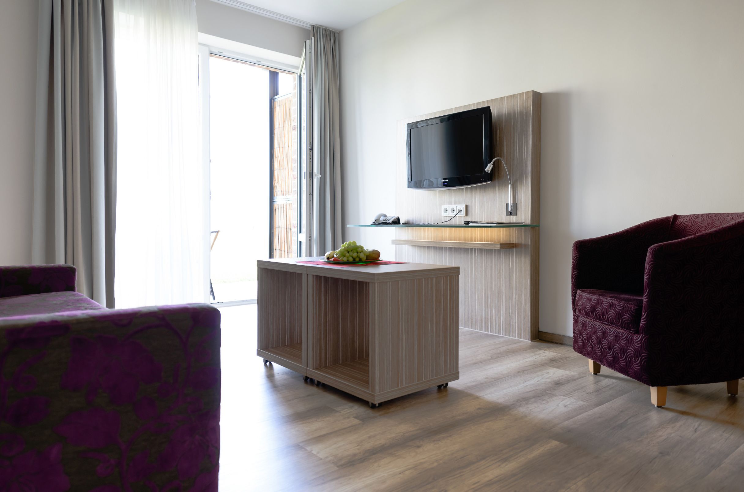 Uma sala de estar moderna com uma televisão de tela plana na parede, um móvel de madeira com tampo de vidro e uma poltrona roxa.