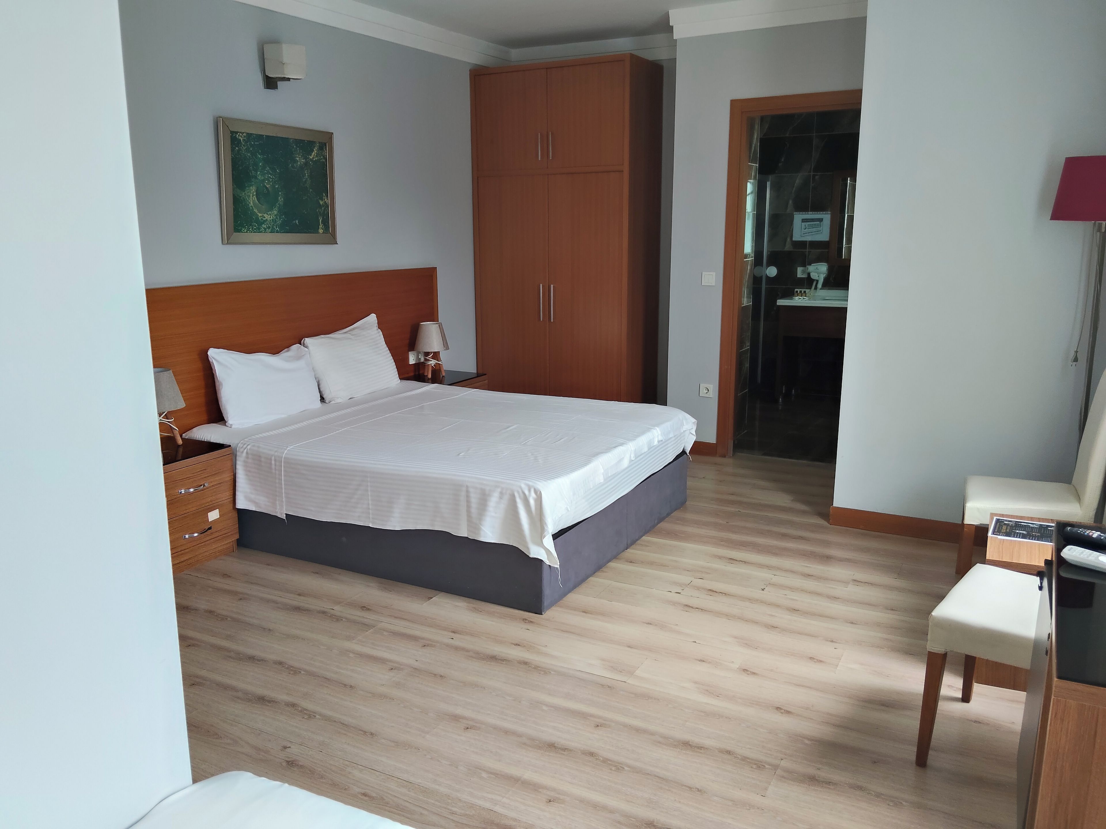 Quarto de hotel com cama, guarda-roupa de madeira, mesa de cabeceira, abajur e escrivaninha com cadeira.