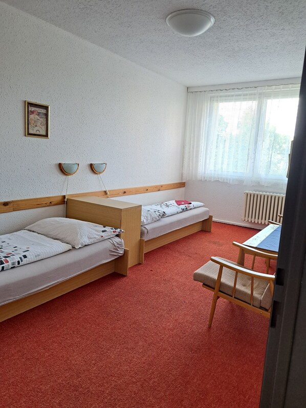 Quarto com duas camas de solteiro, uma escrivaninha de madeira, uma cadeira e janela com cortinas leves.