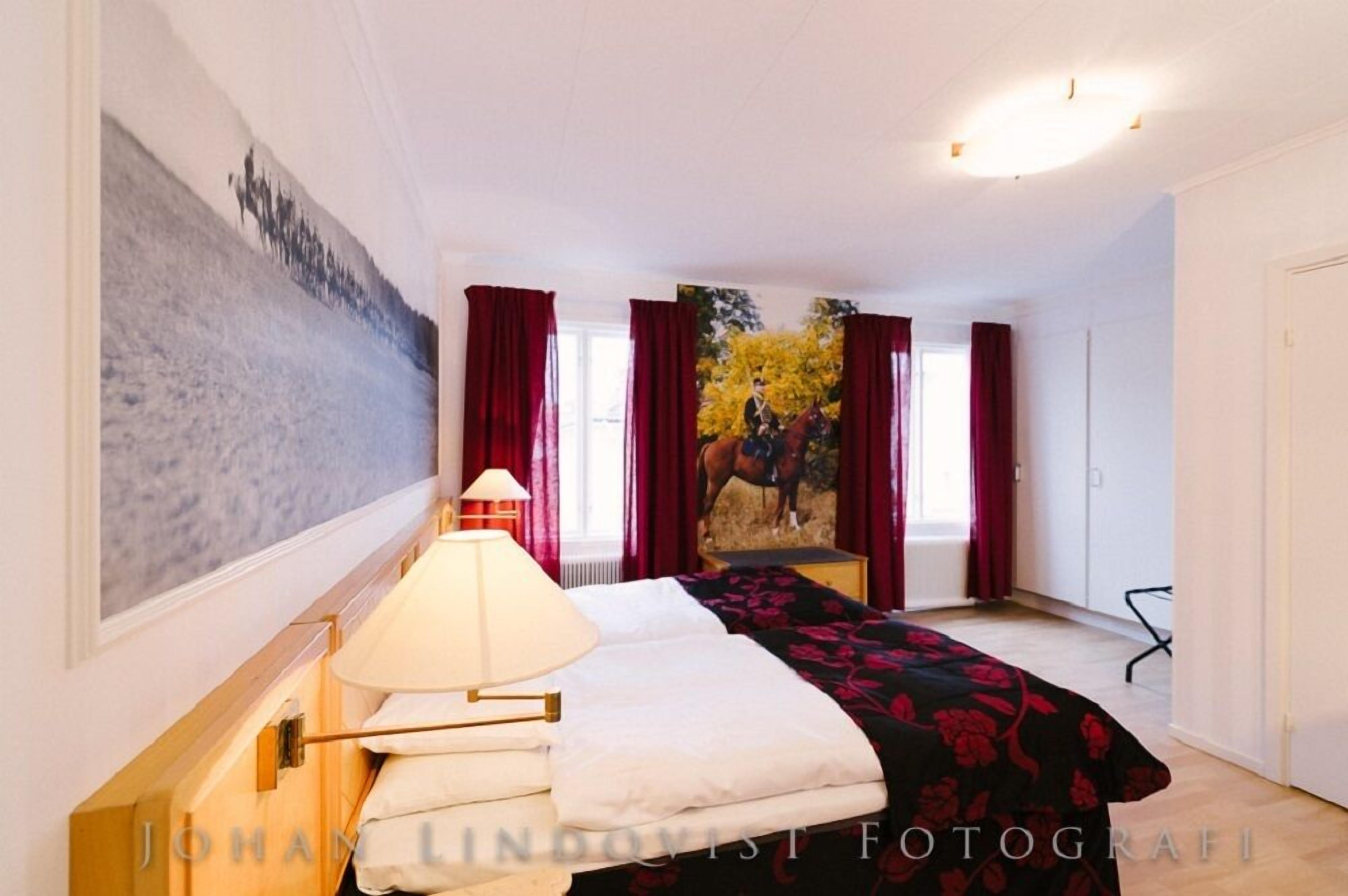 Quarto de hotel com cama, luminária, pintura na parede e janela com cortinas.