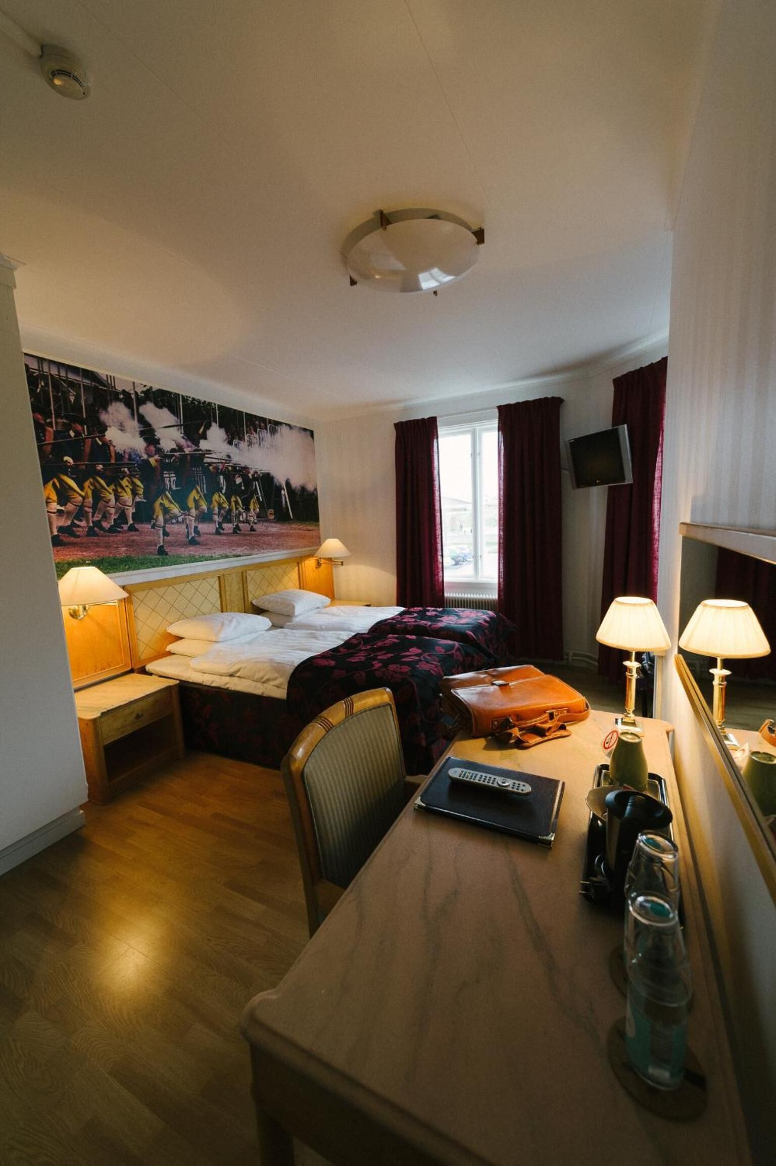 Quarto de hotel com cama, escrivaninha, cadeira, mesa, luminária, mala, televisão e janela com cortinas.