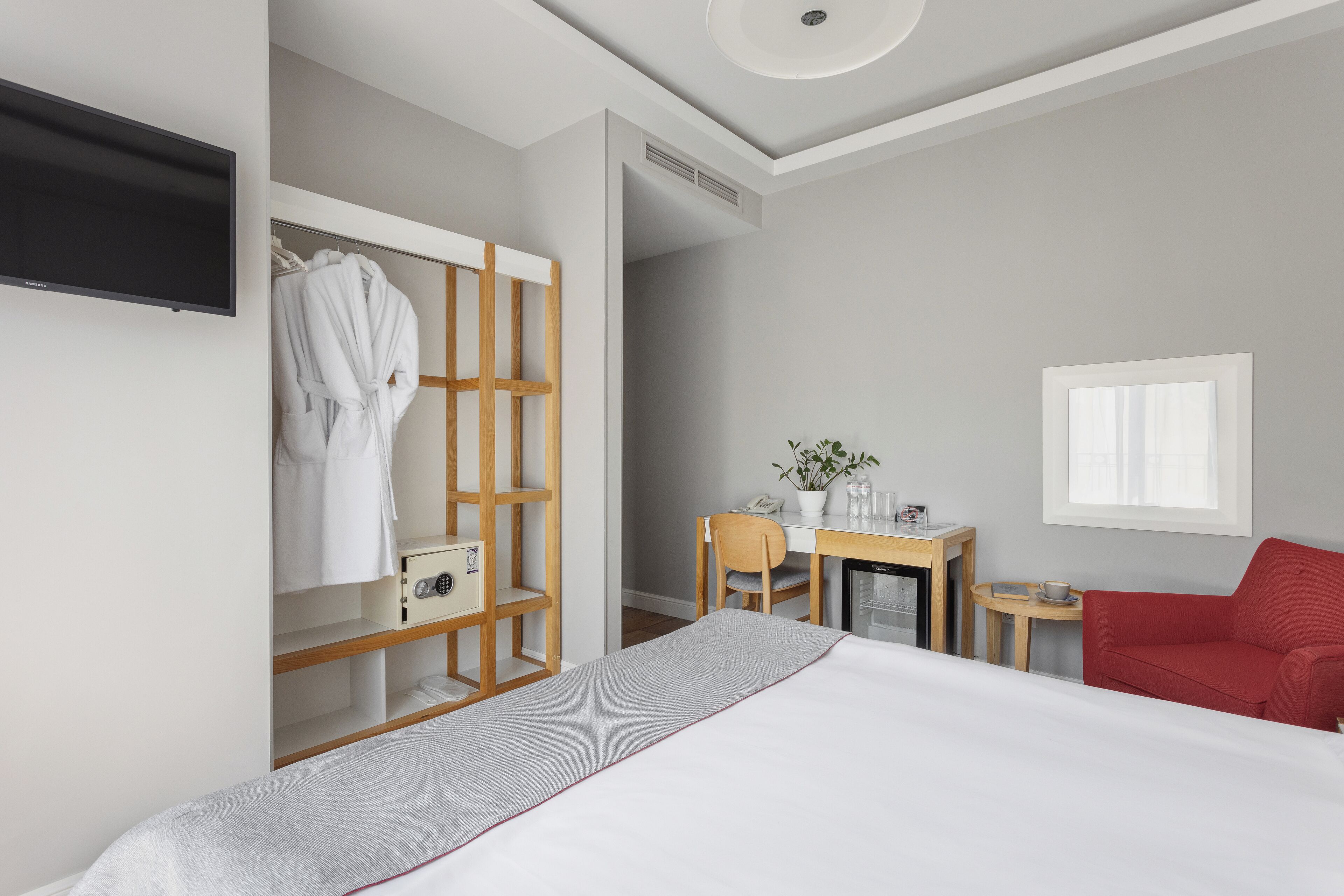 Quarto de hotel com cama, televisão, guarda-roupa de madeira, escrivaninha com cadeira, poltrona vermelha e janela.