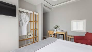 Quarto de hotel com cama, televisão, guarda-roupa de madeira, escrivaninha com cadeira, poltrona vermelha e janela.