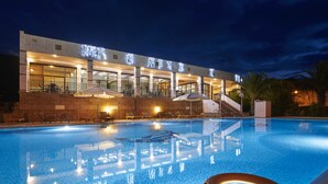Uma piscina com espreguiçadeiras e guarda-sóis, um salva-vidas e um edifício com sinalização iluminada.