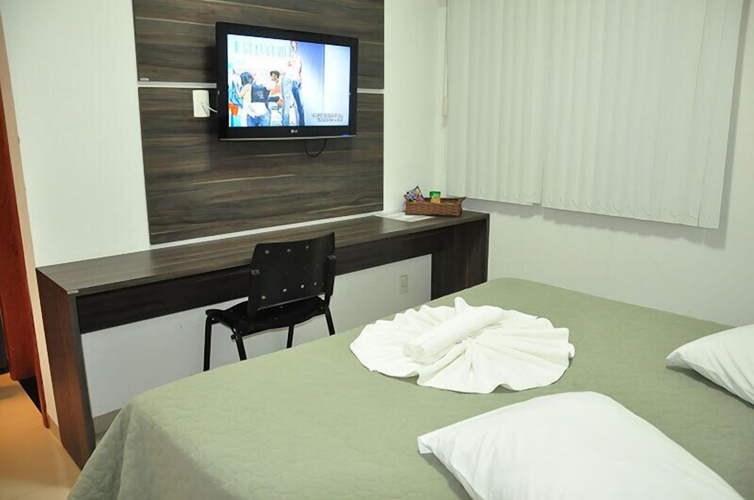 Quarto de hotel com cama, escrivaninha com cadeira, televisão na parede e janela com persianas.