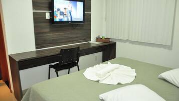 Quarto de hotel com cama, escrivaninha com cadeira, televisão na parede e janela com persianas.