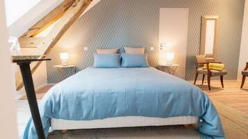 Quarto com teto inclinado, cama com roupa de cama azul, duas luminárias de cabeceira, uma cadeira e um espelho na parede.