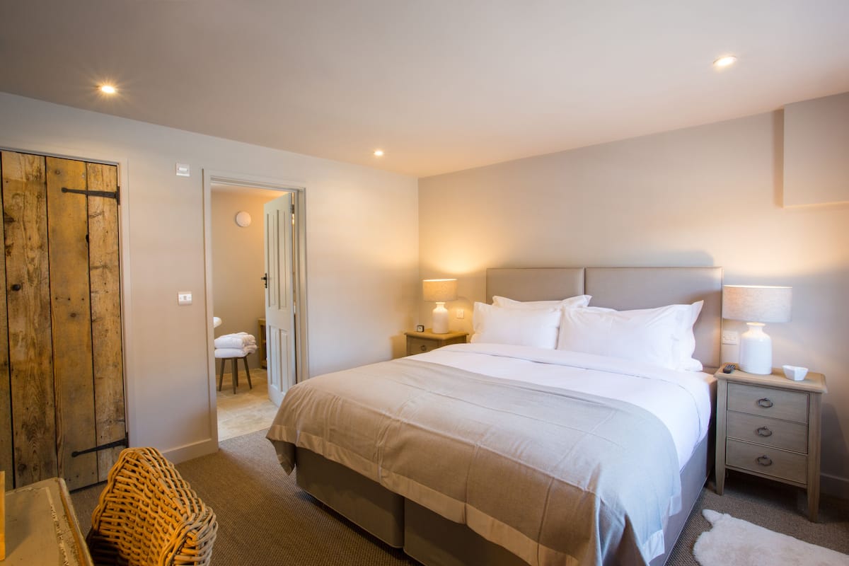 Quarto com uma cama grande, mesas de cabeceira com luminárias, uma porta de madeira e um pequeno espaço de estar.