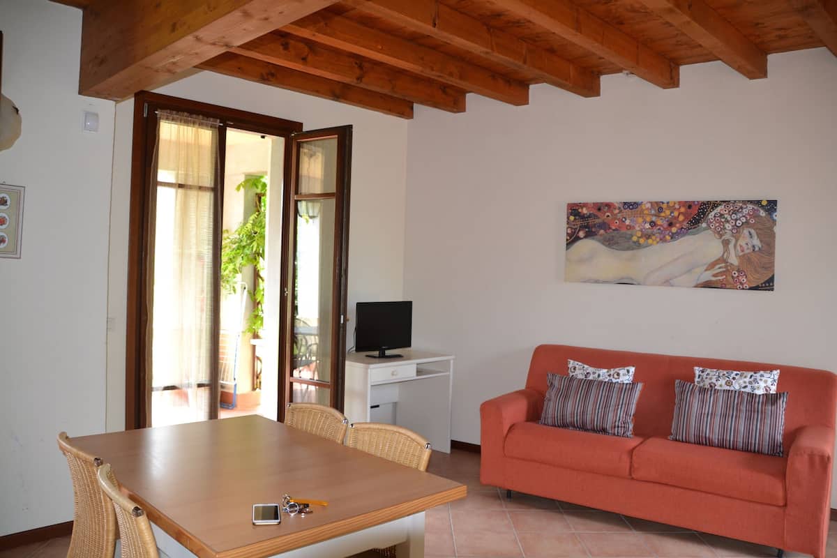 Quarto com mesa de madeira, cadeiras de vime, sofá vermelho com almofadas estampadas, uma televisão sobre uma escrivaninha branca e um quadro na parede.