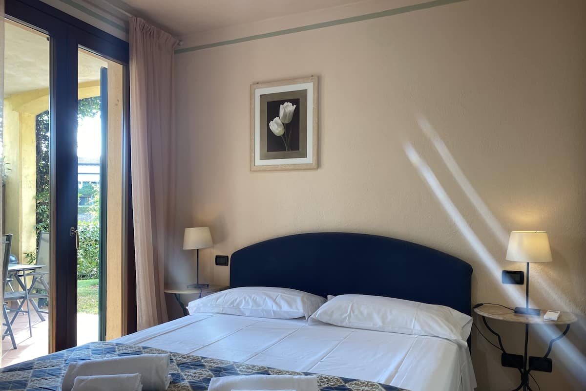 Quarto de hotel com uma cama grande, duas luminárias de cabeceira, um quadro na parede e vista para o exterior por uma porta deslizante.