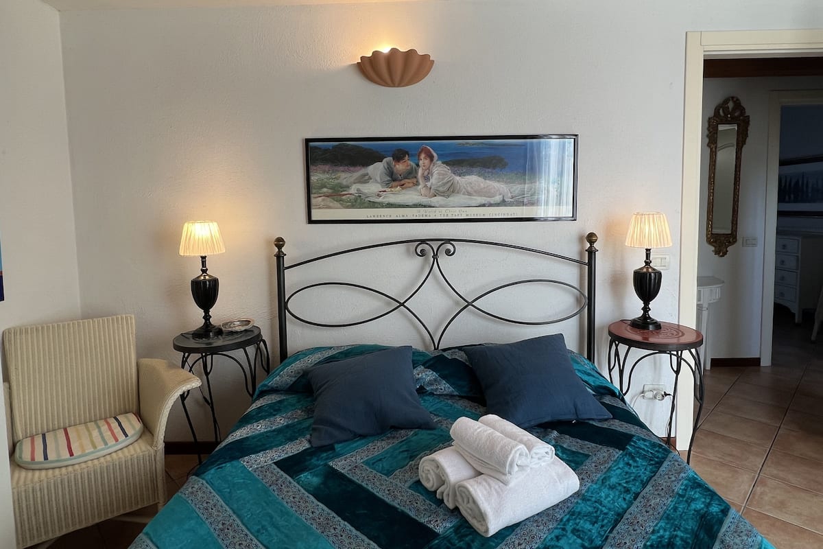 Uma cama bem arrumada com um edredom estampado, duas luminárias de cabeceira, uma cadeira e um quadro na parede.