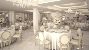 Uma ampla sala de banquetes com várias mesas redondas preparadas para refeições, elegantes lustres e arquitetura clássica.