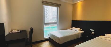 Quarto de hotel com duas camas, uma escrivaninha, uma cadeira, uma janela com vista para edifícios e um telefone na mesa de cabeceira.