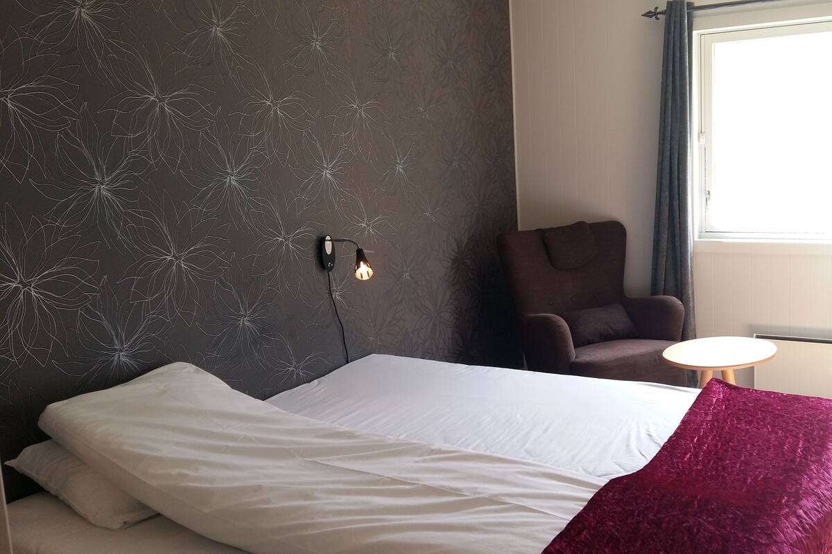 Quarto com cama, cadeira, mesa e janela com cortinas.