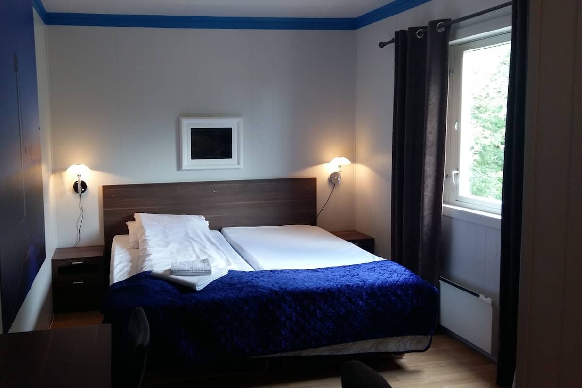 Uma cama de solteiro com um cobertor azul, duas luminárias de cabeceira, uma mesinha de cabeceira pequena e uma janela com cortinas.