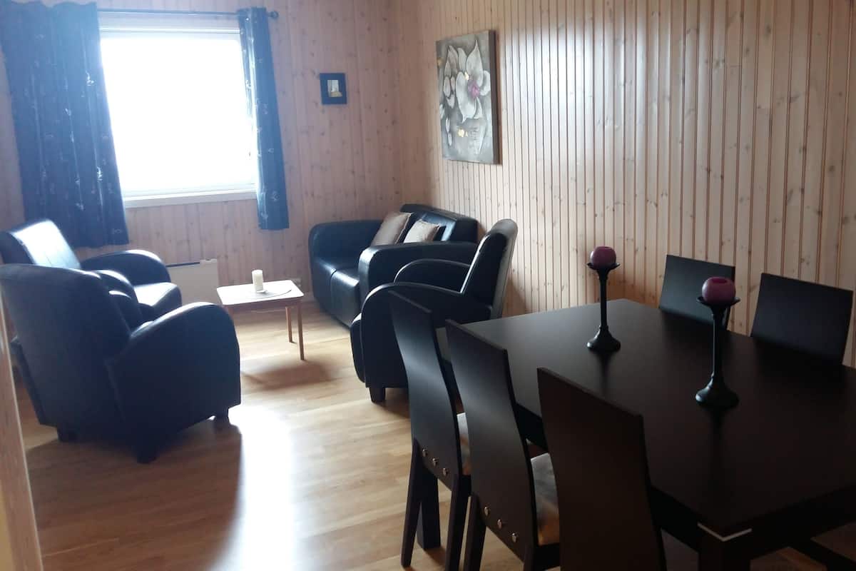 Um quarto com parede revestida de madeira, um sofá de couro preto, uma mesa de jantar com cadeiras e uma janela com cortinas.