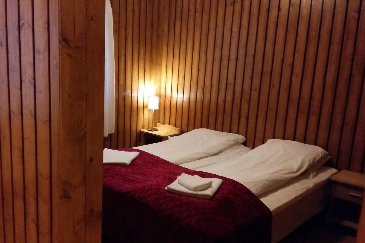 Quarto com painéis de madeira, duas camas, uma mesa de cabeceira e uma luminária fixada na parede.