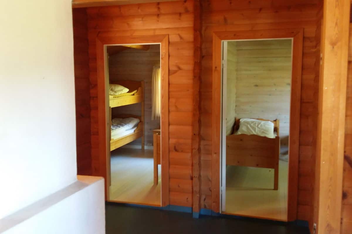 Uma cabana de madeira com duas beliches, uma delas com colchão e travesseiros à mostra, e uma porta que dá para outro quarto.