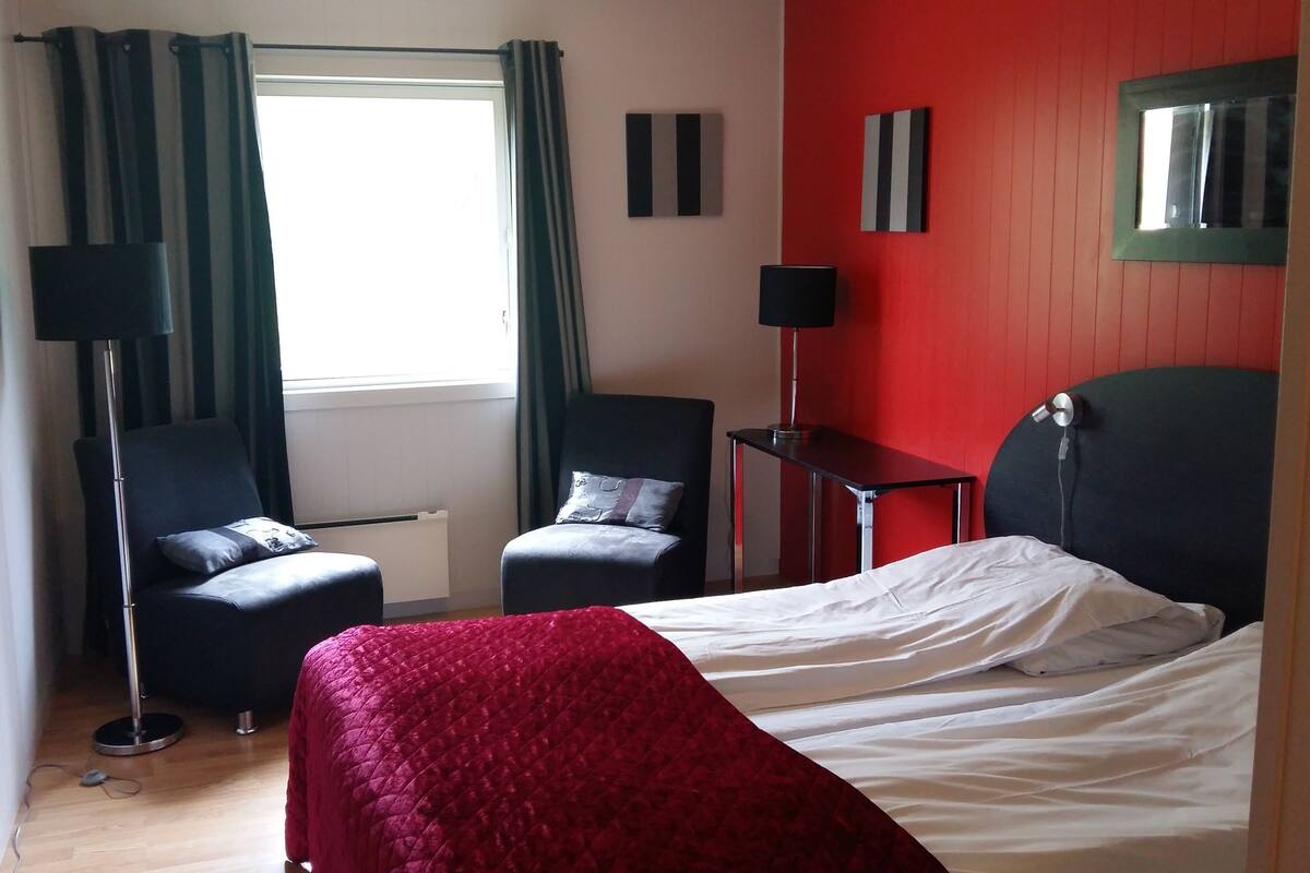 Quarto de hotel com cama, duas cadeiras, escrivaninha, luminária de chão e janela com cortinas.