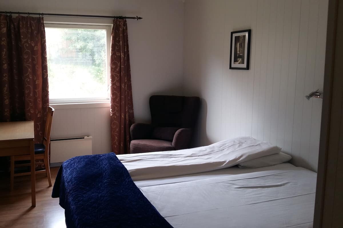 Quarto pequeno com uma cama de solteiro, escrivaninha e cadeira, janela com cortinas e um quadro na parede.