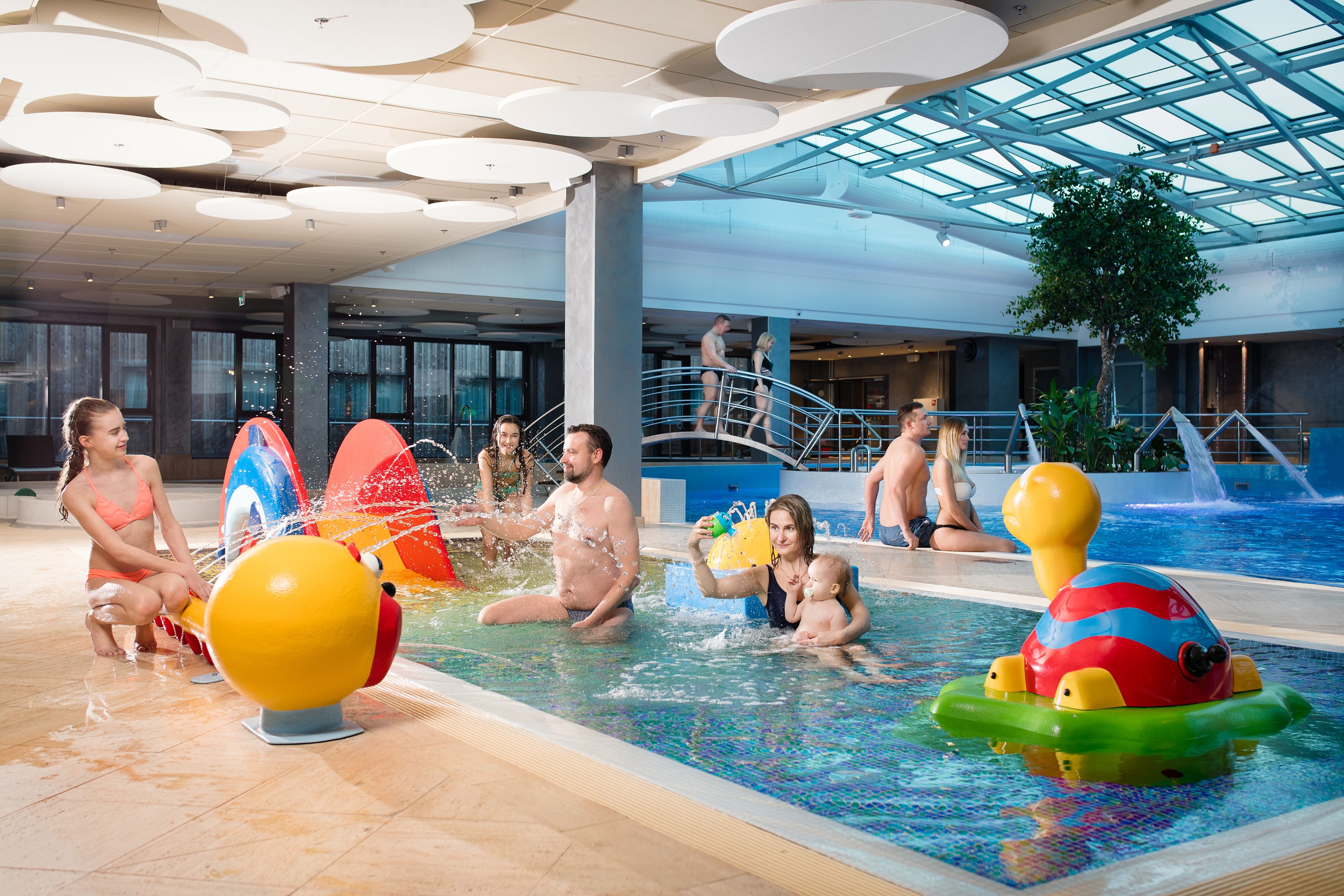 Área interna de piscina com brinquedos aquáticos, teto de vidro e escada.