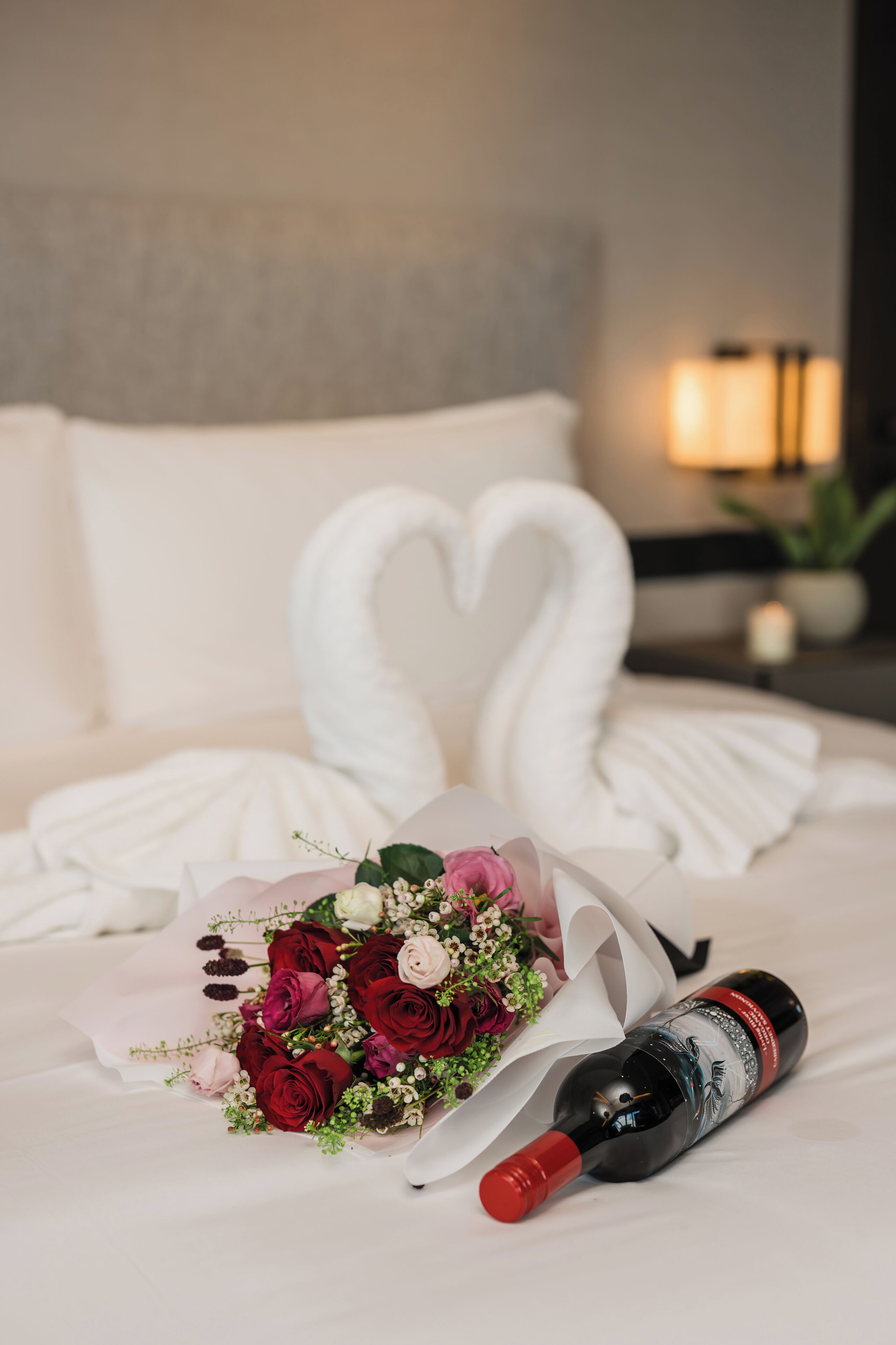 Uma cama com travesseiros em formato de coração, um buquê de flores e uma garrafa de vinho.