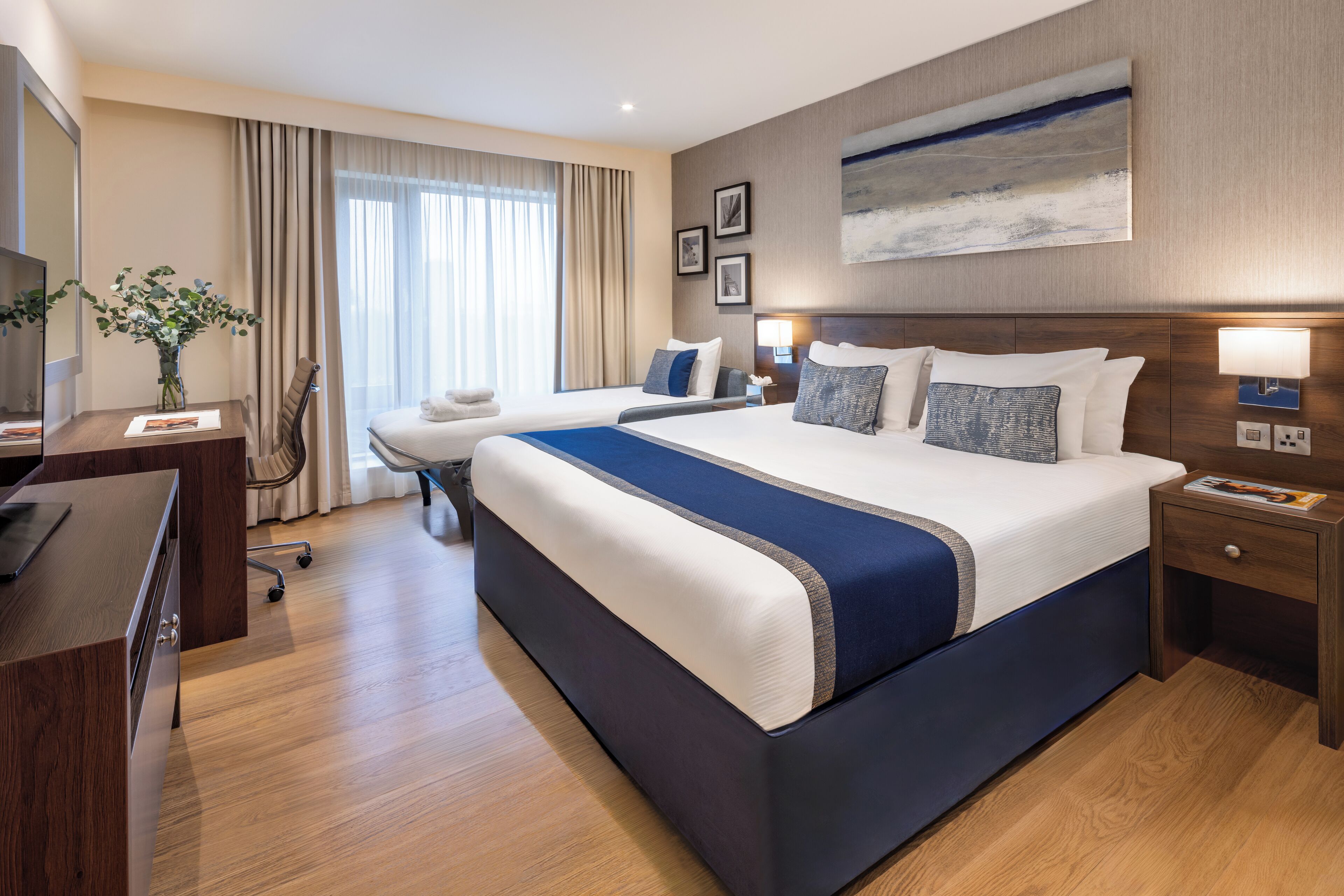 Quarto de hotel com uma cama grande, uma mesa e uma cadeira. Há uma janela com cortinas, um quadro na parede e um abajur na mesinha de cabeceira.