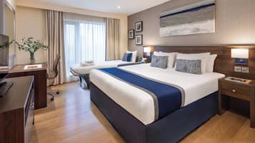 Quarto de hotel com uma cama grande, uma mesa e uma cadeira. Há uma janela com cortinas, um quadro na parede e um abajur na mesinha de cabeceira.