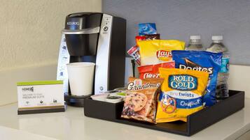 Espaço de café da manhã em quarto de hotel, com cafeteira Keurig, uma xícara, lanches variados e um sinal de boas-vindas.