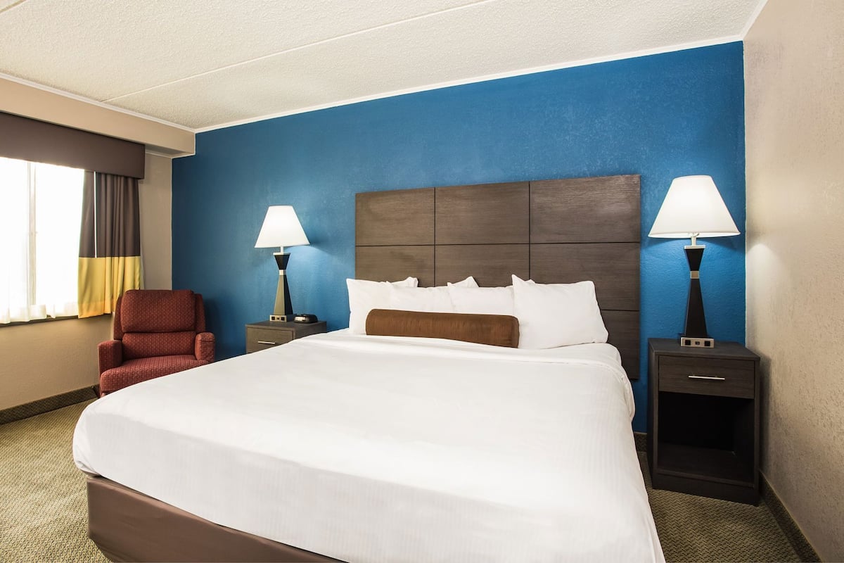 Quarto de hotel com uma cama grande, duas mesas de cabeceira com abajures, uma poltrona vermelha e uma janela com cortinas.