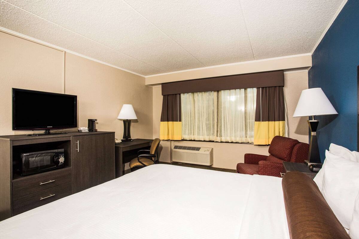 Quarto de hotel com uma cama, uma escrivaninha, uma televisão, uma cadeira e um abajur.