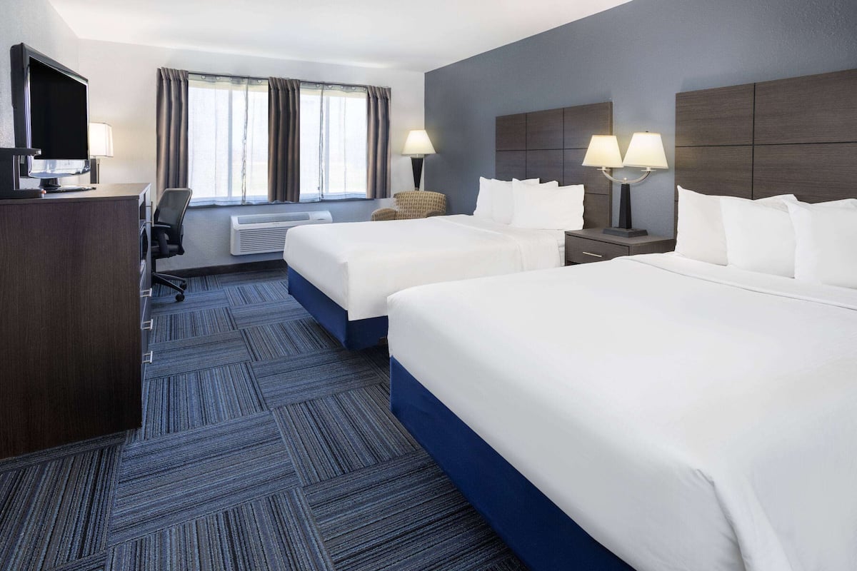 Quarto de hotel com duas camas, uma mesa de trabalho com uma TV, uma cadeira, uma janela com cortinas e duas luminárias de cabeceira.