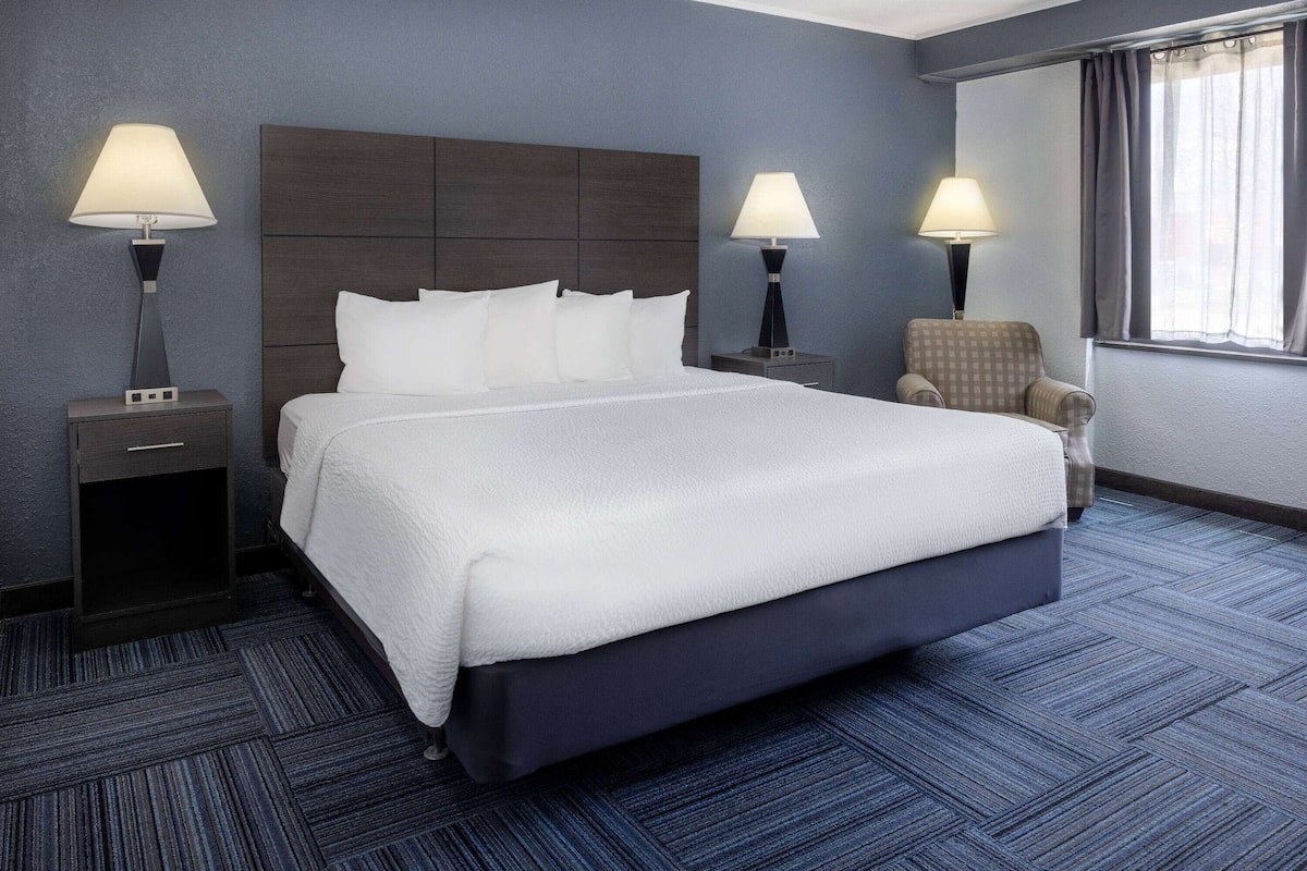 Quarto de hotel com uma cama grande, duas mesas de cabeceira com luminárias, uma cadeira e uma janela com cortinas.