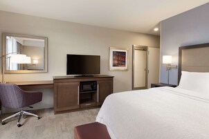 Quarto de hotel com uma cama, escrivaninha com televisão, cadeira, micro-ondas, quadro e uma porta.