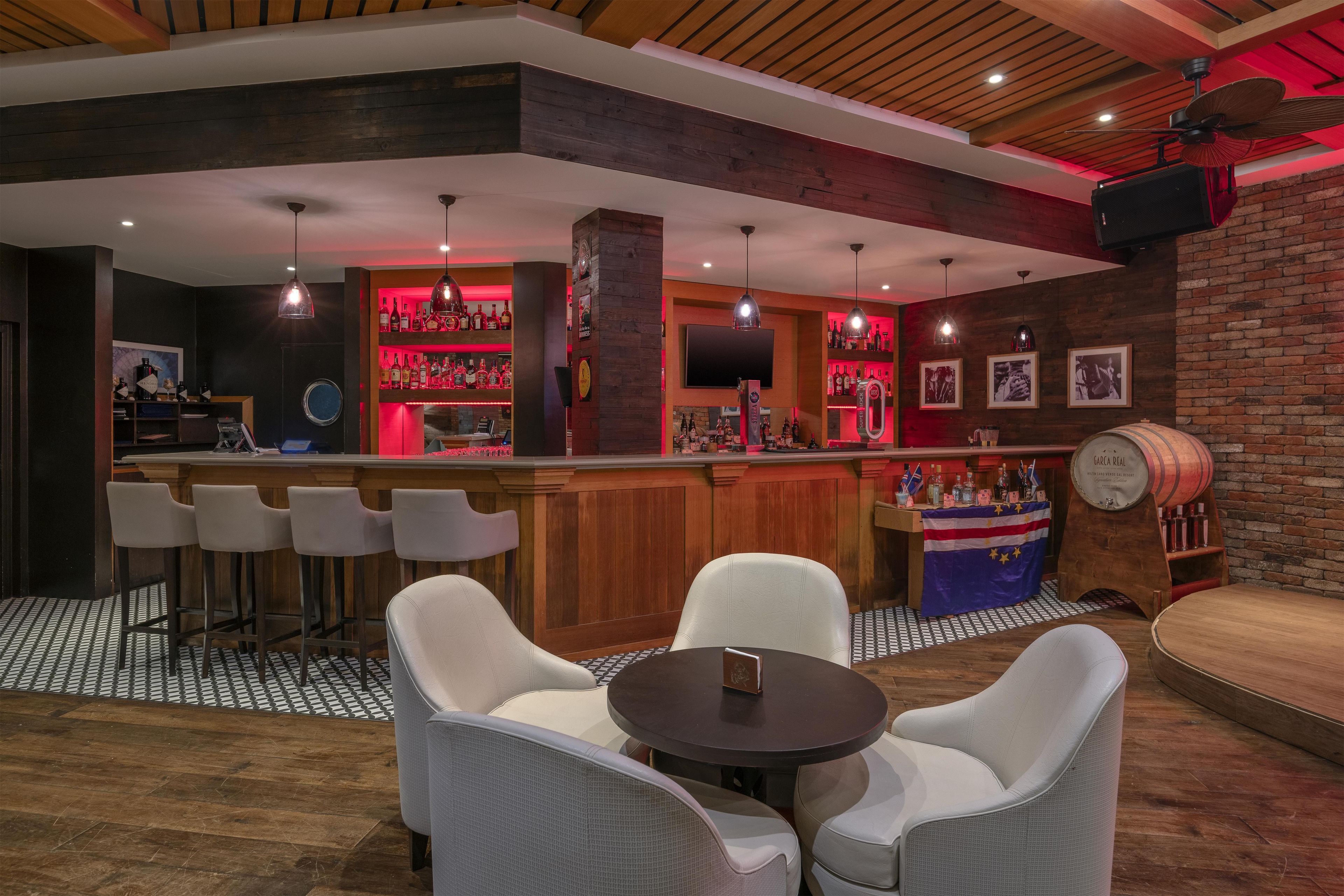 Uma área de bar com teto de madeira, banquetas, uma mesa redonda e balcão com garrafas.