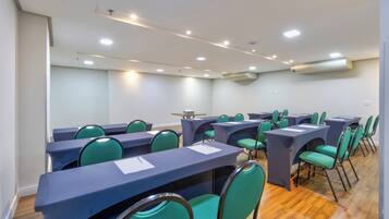 Uma sala de conferência com mesas e cadeiras dispostas em fileiras, luminárias no teto e um aparelho de ar condicionado.