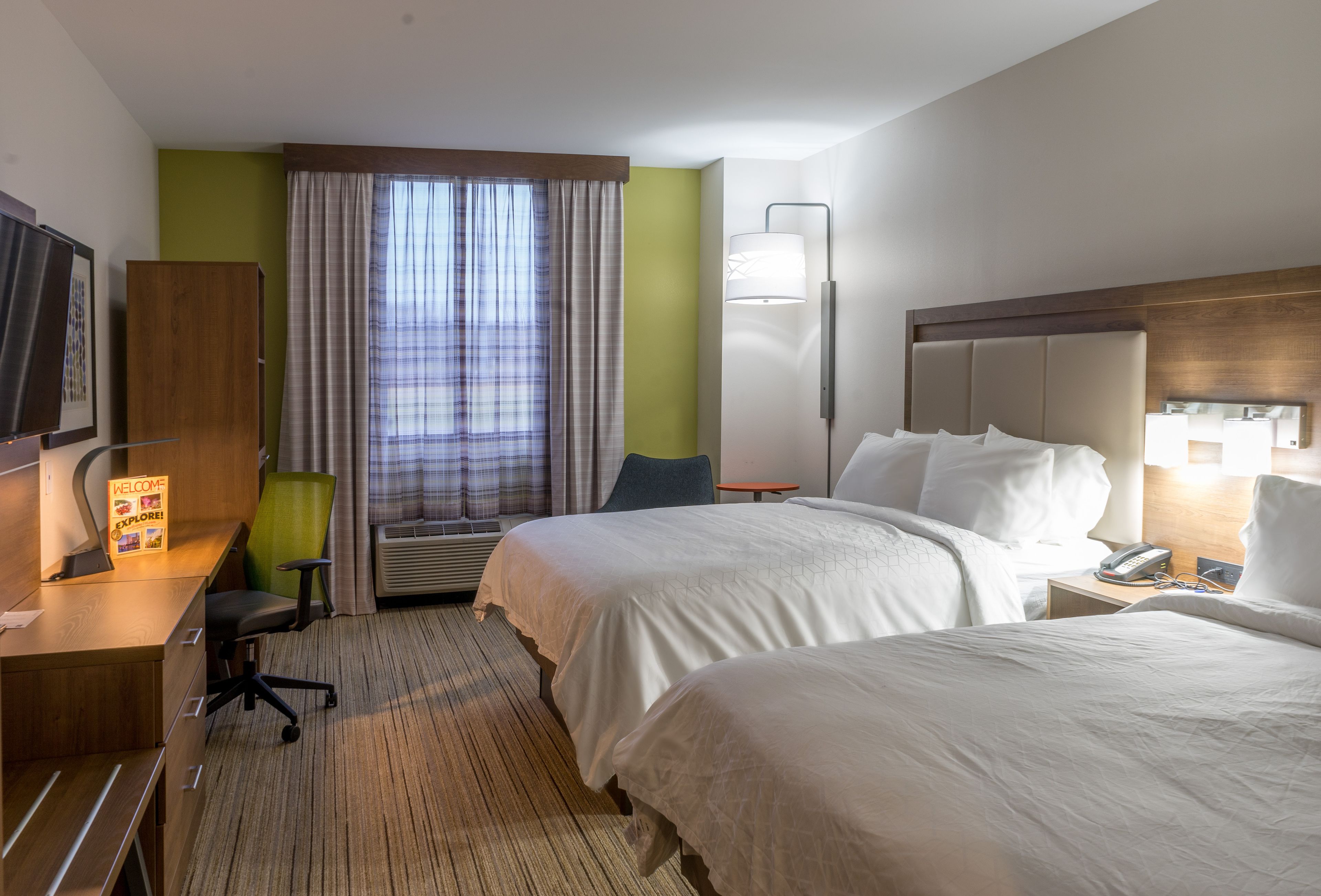 Quarto de hotel com duas camas, uma escrivaninha com cadeira, televisão, luminária e janela com cortinas.