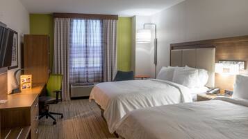 Quarto de hotel com duas camas, uma escrivaninha com cadeira, televisão, luminária e janela com cortinas.