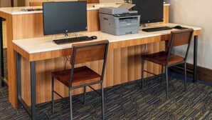 Quarto de hotel com escrivaninha de madeira, dois monitores de computador, uma impressora e duas cadeiras.