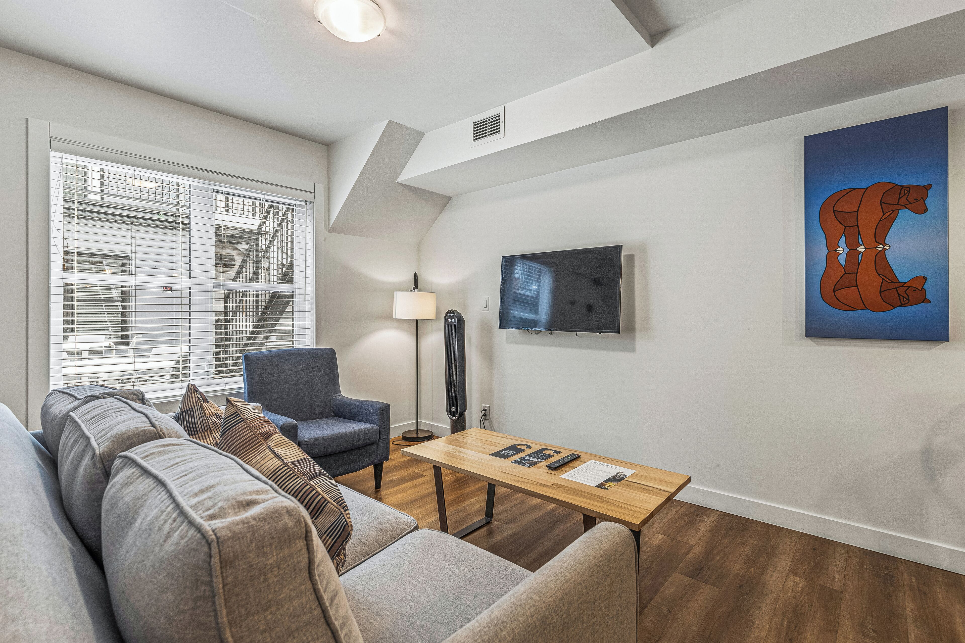 Uma sala de estar moderna com sofá, poltrona, luminária de chão e uma mesa com controles remotos.